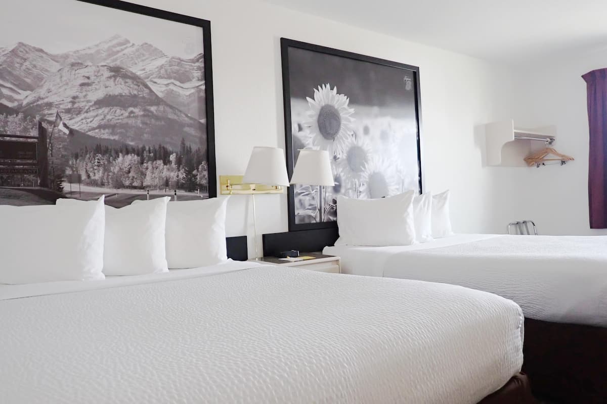 Quarto de hotel com duas camas, roupa de cama branca, mesa de cabeceira com luminárias e duas grandes molduras na parede.