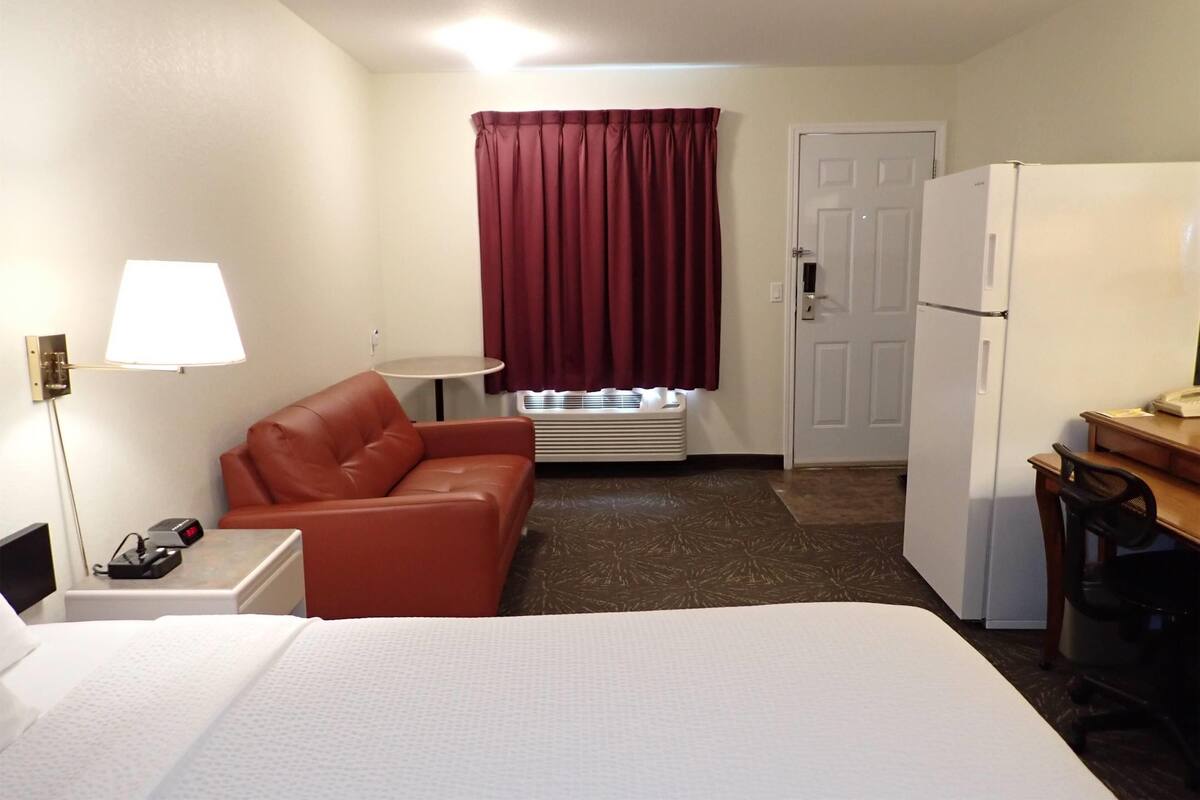 Quarto de hotel com cama, uma poltrona de couro vermelha, uma mesinha de cabeceira com abajur, um refrigerador e uma janela com cortinas.
