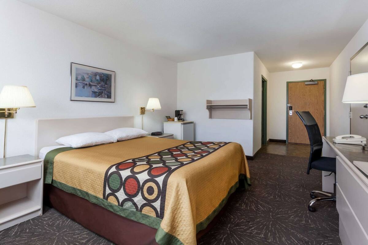 Quarto de hotel com cama, abajures, escrivaninha com cadeira, telefone e porta.