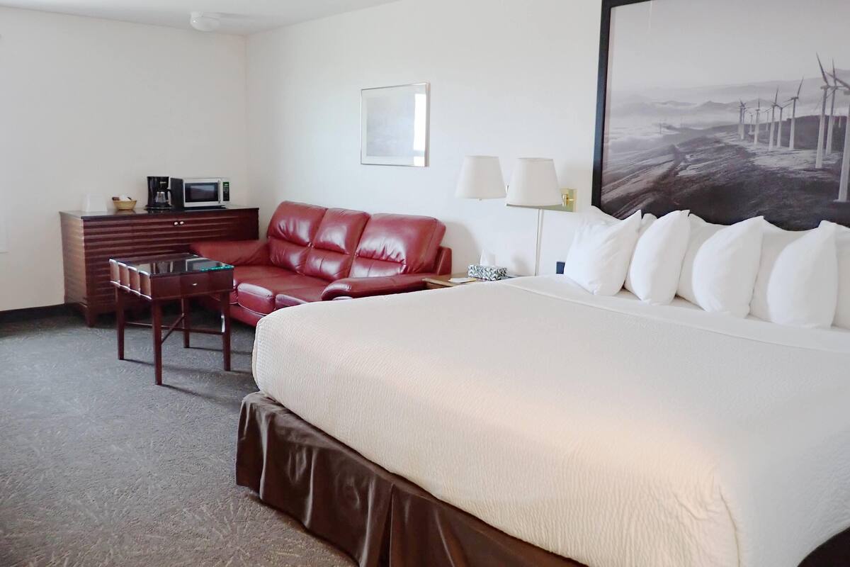 Quarto de hotel com uma cama grande, um sofá de couro vermelho, uma escrivaninha de madeira com micro-ondas e um quadro na parede.