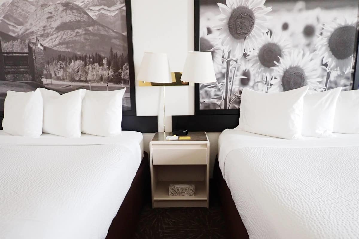 Quarto de hotel com duas camas, uma mesa de cabeceira com abajur, uma mesinha e um quadro na parede com uma paisagem de montanhas e girassóis.