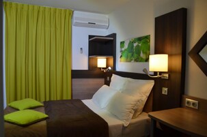 Quarto de hotel com uma cama, cortinas verdes, ar condicionado fixado na parede e duas luminárias de cabeceira.