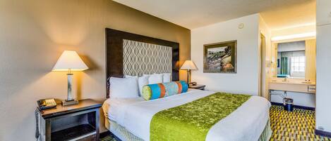 Quarto de hotel com uma cama grande, duas luminárias laterais, uma mesa de cabeceira, um quadro na parede e banheiro com pia e espelho.
