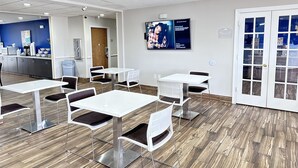 Uma sala de jantar moderna com mesas e cadeiras brancas, cozinha ao fundo e um pôster grande na parede.