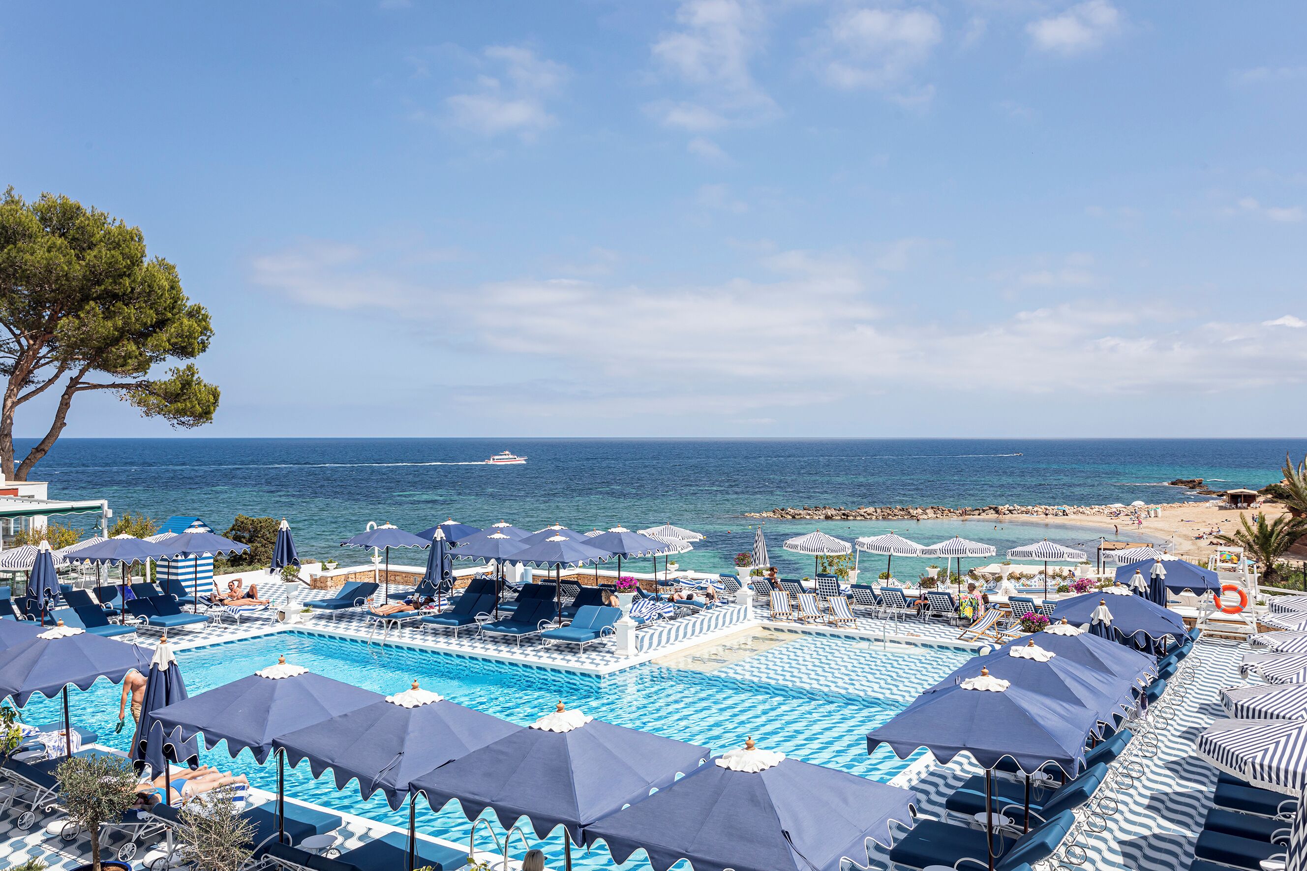 Uma área à beira da piscina com espreguiçadeiras e guarda-sóis, vista desobstruída para o mar e uma praia de areia ao fundo.