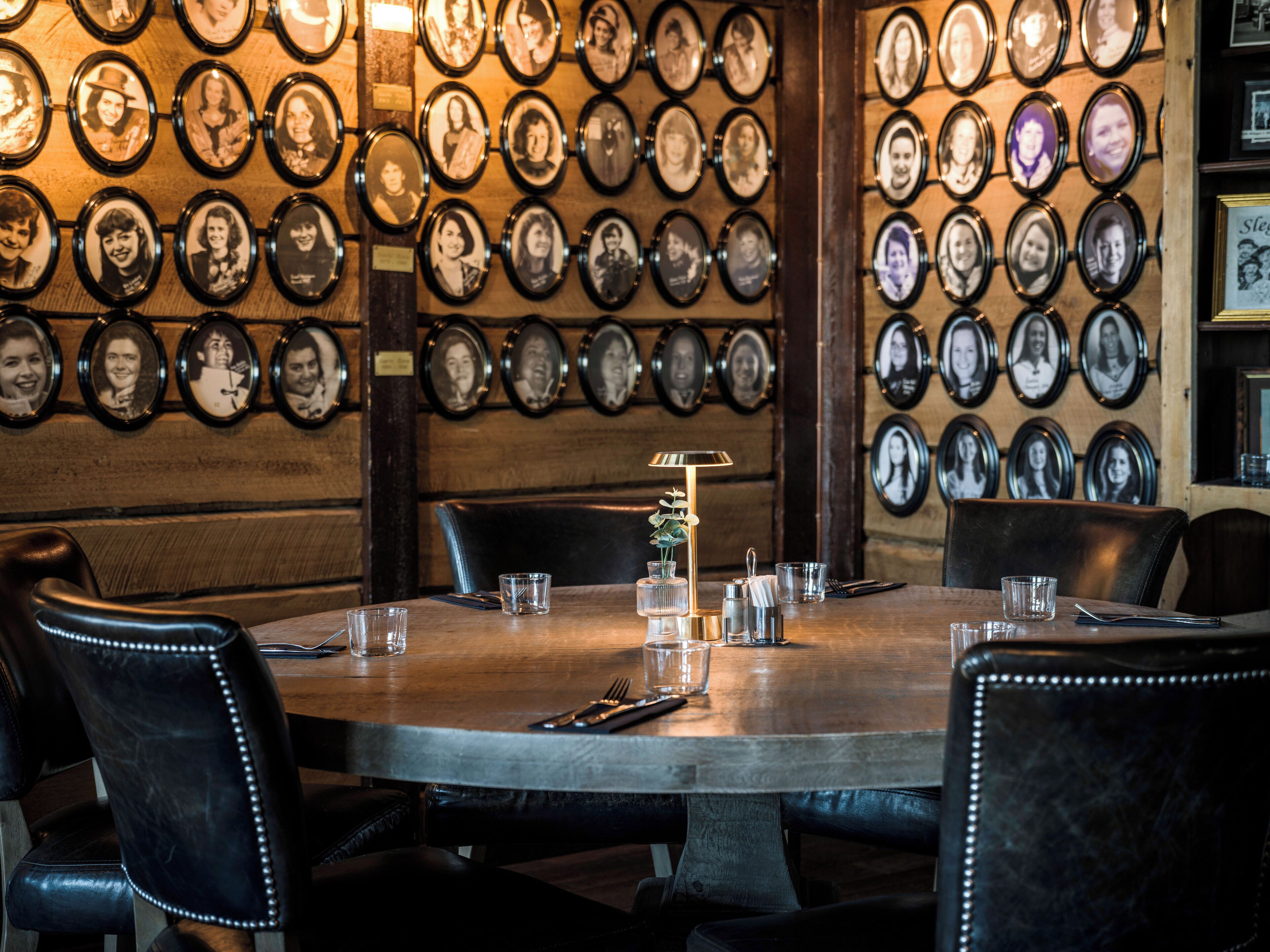 Um restaurante com uma parede de fotos emolduradas, uma mesa redonda com cadeiras e uma pequena mesa com abajur e copos.