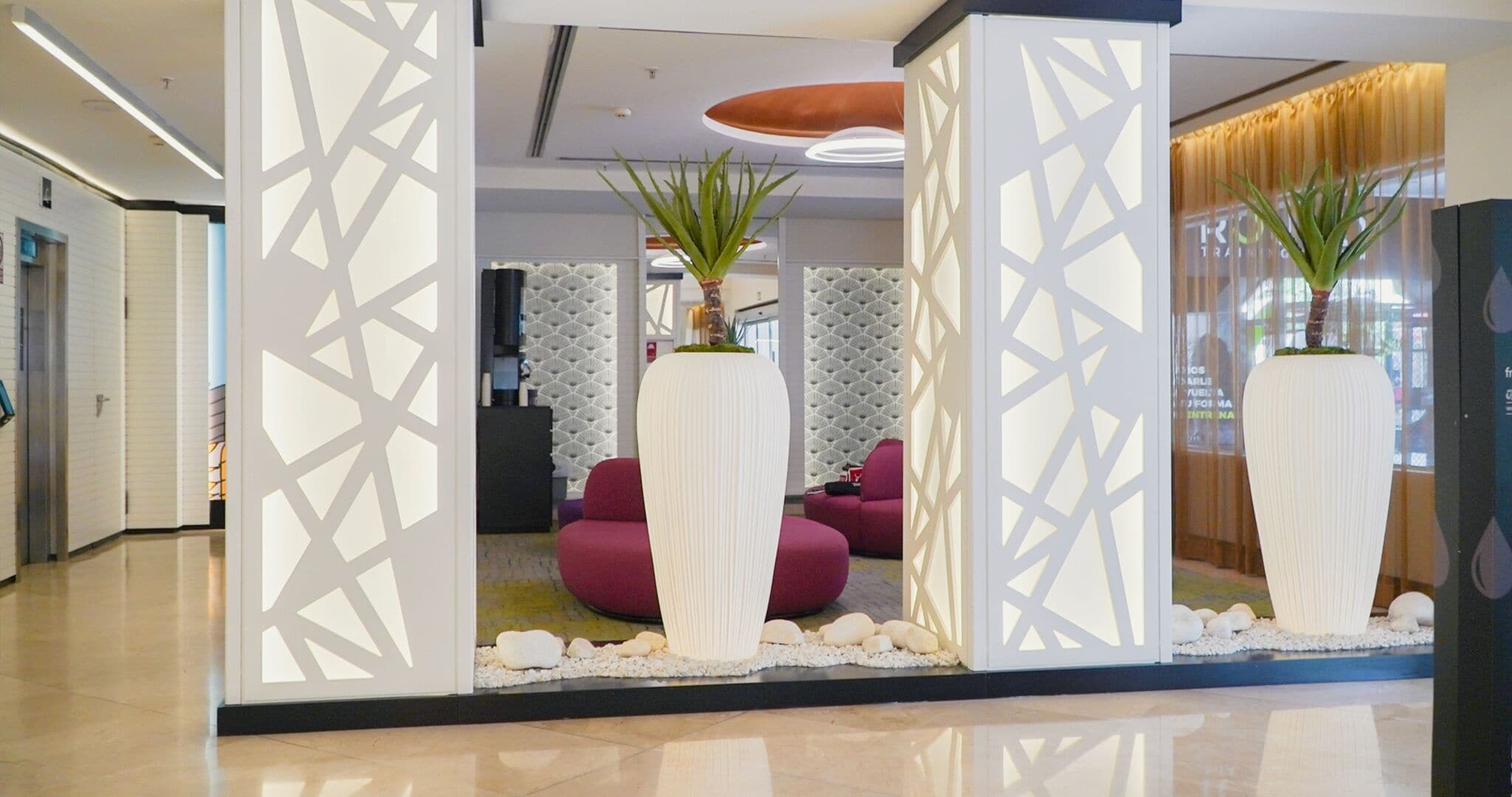 Um lobby moderno com divisória geométrica, balcão de recepção, sofá vermelho e dois grandes vasos brancos com plantas verdes.