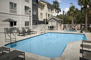 Piscina com espreguiçadeiras e guarda-sóis, cercada por uma cerca branca, com um edifício ao fundo.