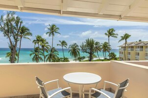 Varanda com mesa e cadeiras brancas, vista para uma praia com palmeiras e céu azul límpido.
