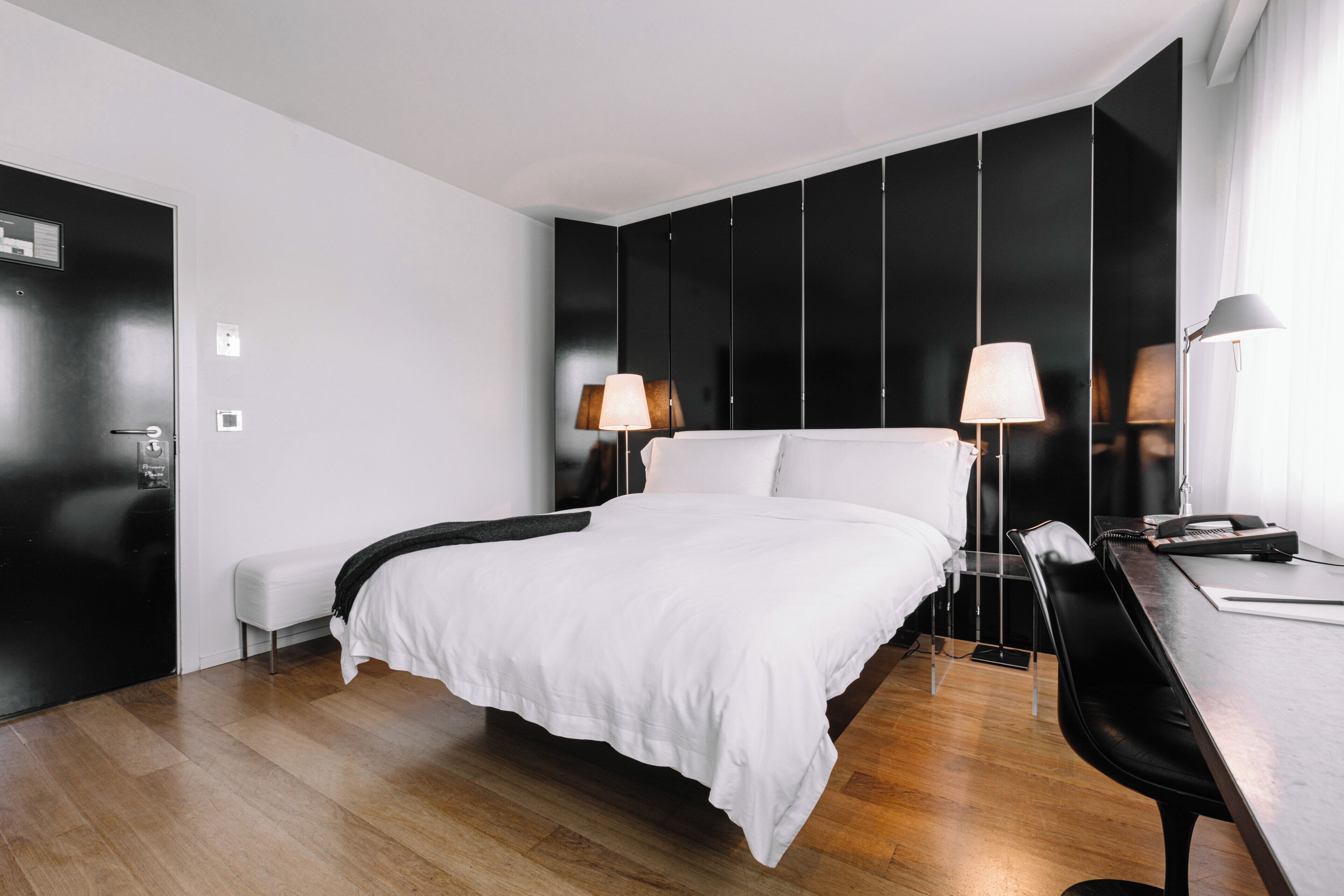 Quarto de hotel moderno com uma cama grande, duas luminárias de cabeceira, uma escrivaninha com cadeira e um guarda-roupa preto.