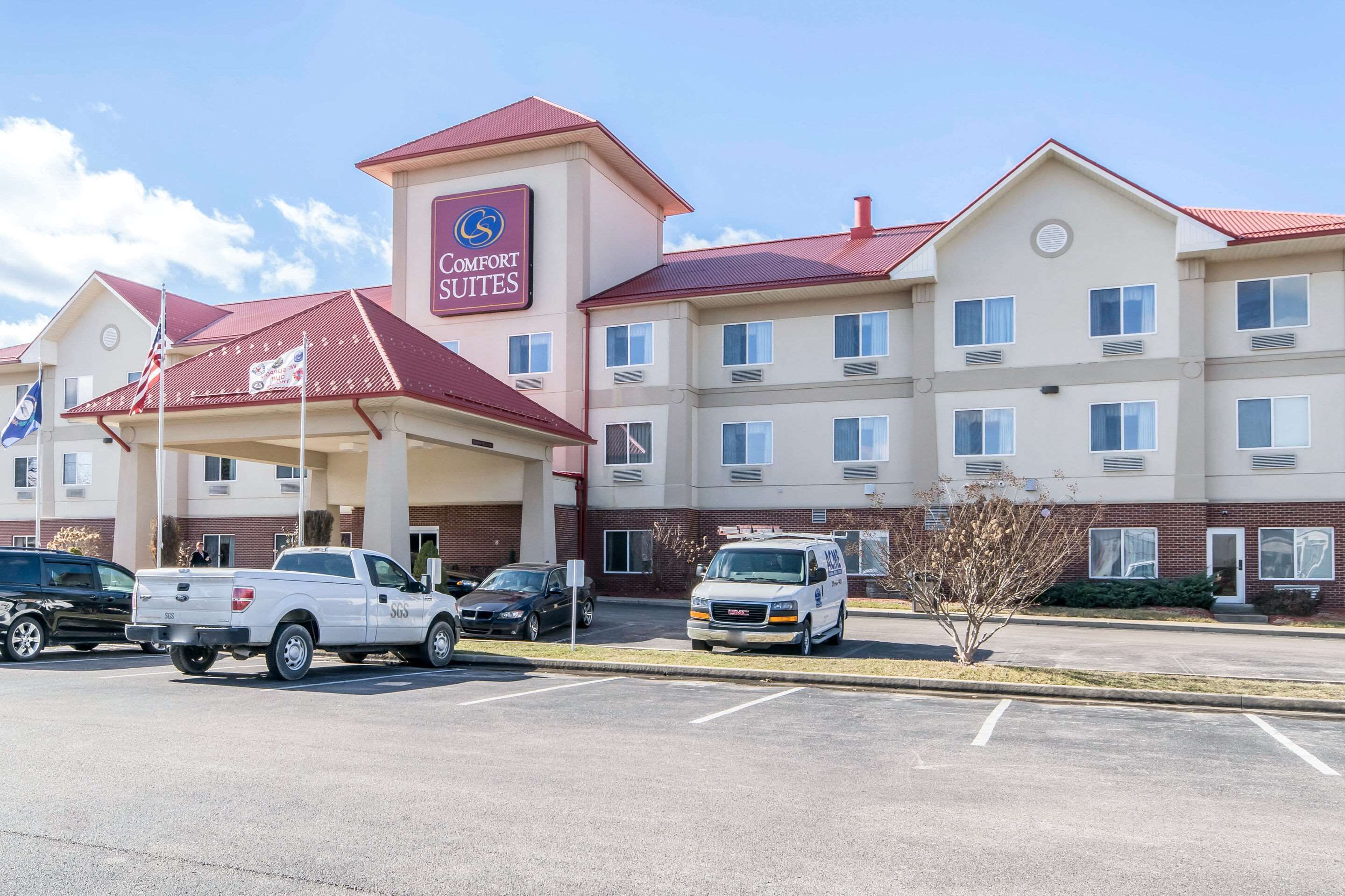 Um hotel Comfort Suites com telhado vermelho e fachada bege. Há várias vagas de estacionamento e uma van branca estacionada na frente.