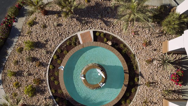 Uma piscina circular com uma fonte central, rodeada por um leito de pedras e palmeiras.