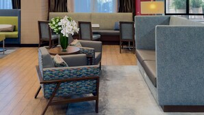 Um saguão de hotel moderno com área de estar, uma janela grande com cortinas e um vaso decorativo com flores.