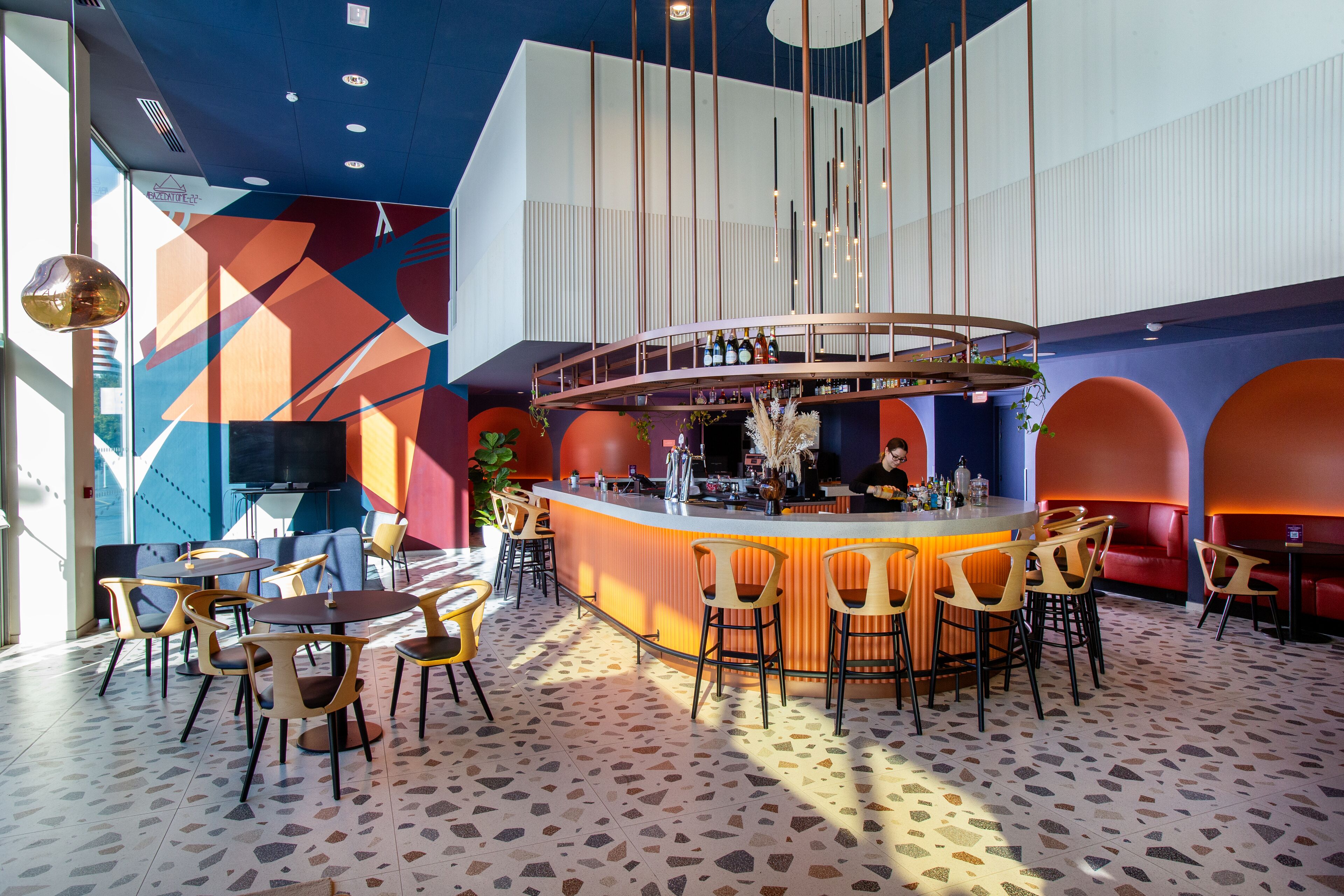 Uma área de bar moderna com balcão central, diversas opções de assento e um design vibrante na parede com motivos geométricos.