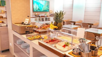 Espaço de buffet com diversas opções de comida, uma televisão na parede e uma área de estar com mesas e cadeiras.