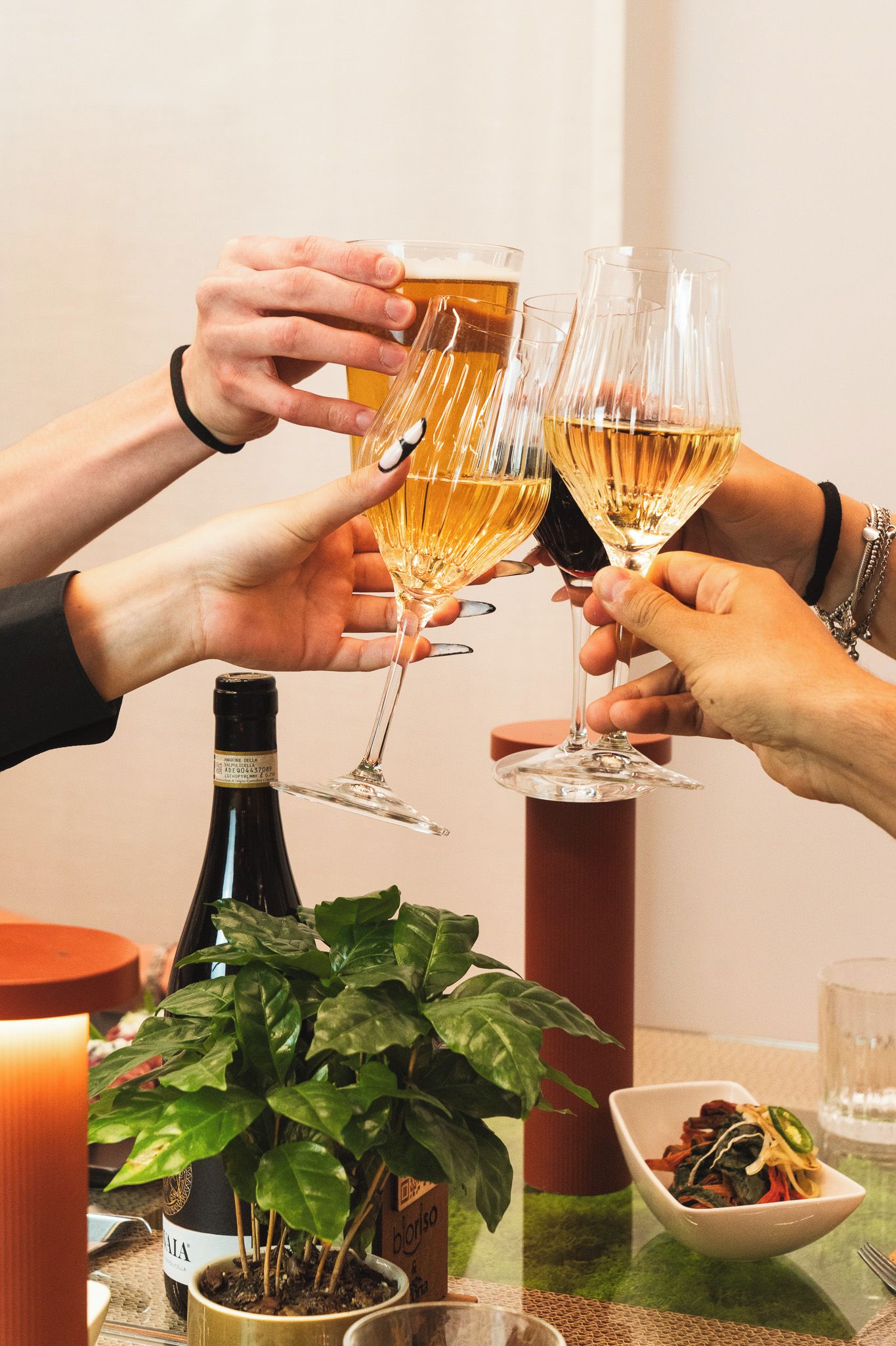 Um grupo de pessoas brindando com copos de vinho, uma garrafa de vinho, uma vela e uma tigela de petiscos em cima de uma mesa.