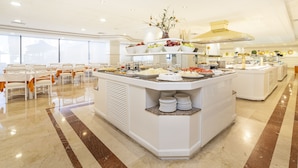 Uma área ampla de buffet com mesas e cadeiras brancas, um balcão central com diversos tipos de comida, e uma grande janela que proporciona iluminação natural.