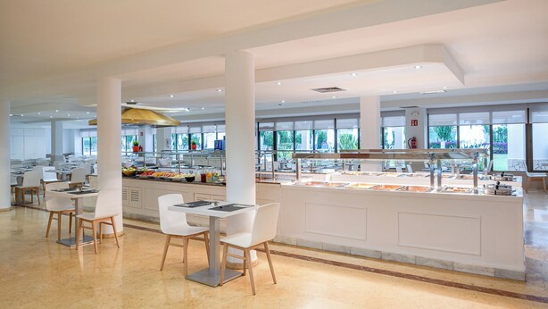 Uma área de refeições moderna com mobiliário branco, um buffet com diversas opções de comida e grandes janelas que oferecem iluminação natural.