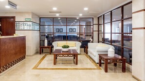 Uma área de recepção com balcão de madeira, sofá branco, mesa de centro e uma janela grande com vista para o interior.