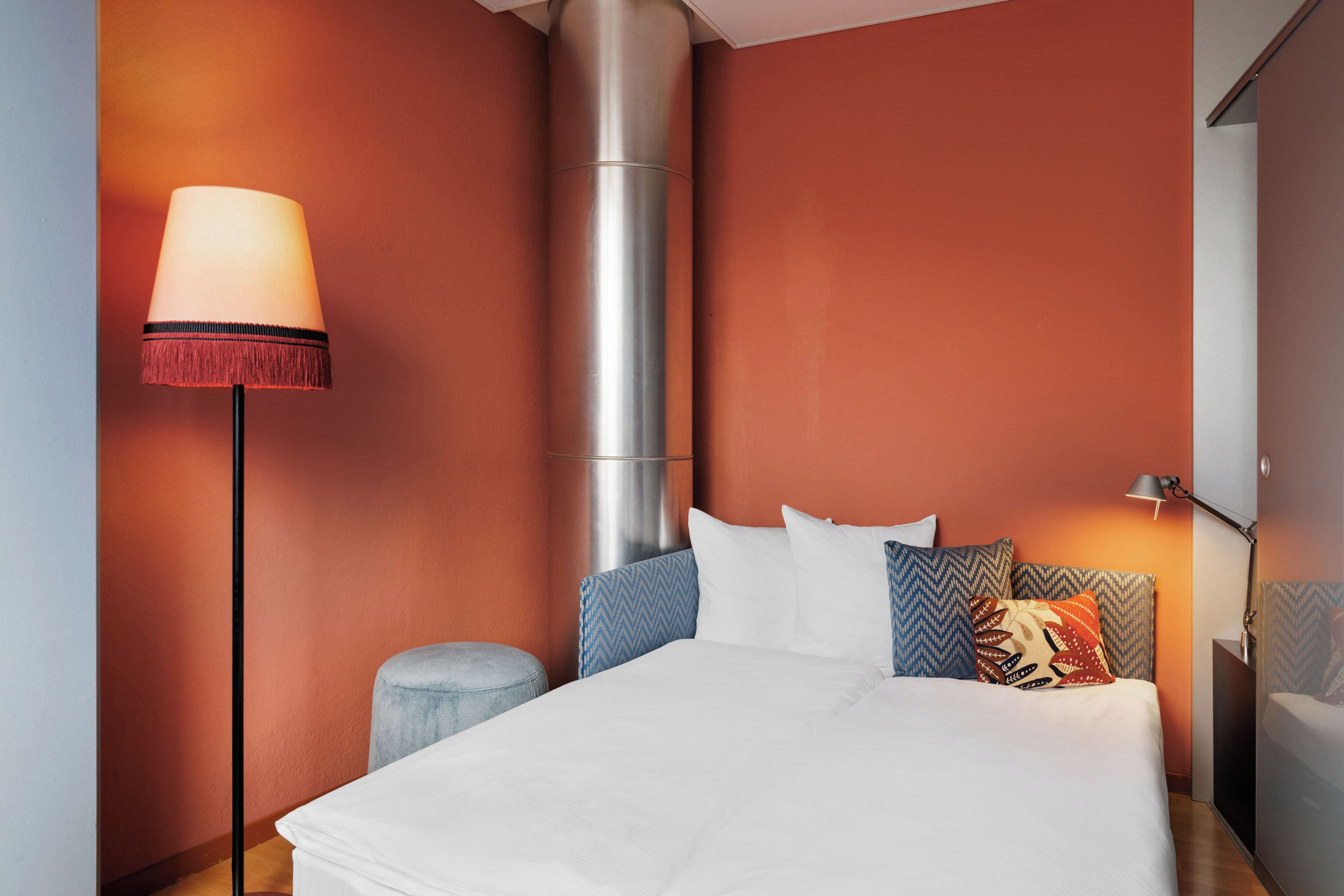 Quarto de hotel com cama, luminária, um registo de ar cilíndrico e espelho fixado na parede.