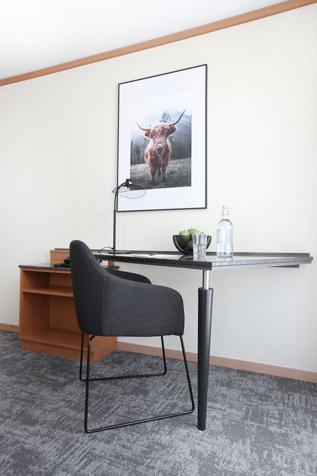 Uma mesa de escritório moderna com um quadro de uma vaca das Terras Altas, um copo de água e uma maçã verde sobre a mesa.