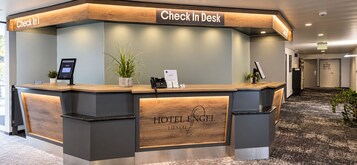 Bancada de recepção de hotel com placa de 'Check In', um display digital e uma planta.