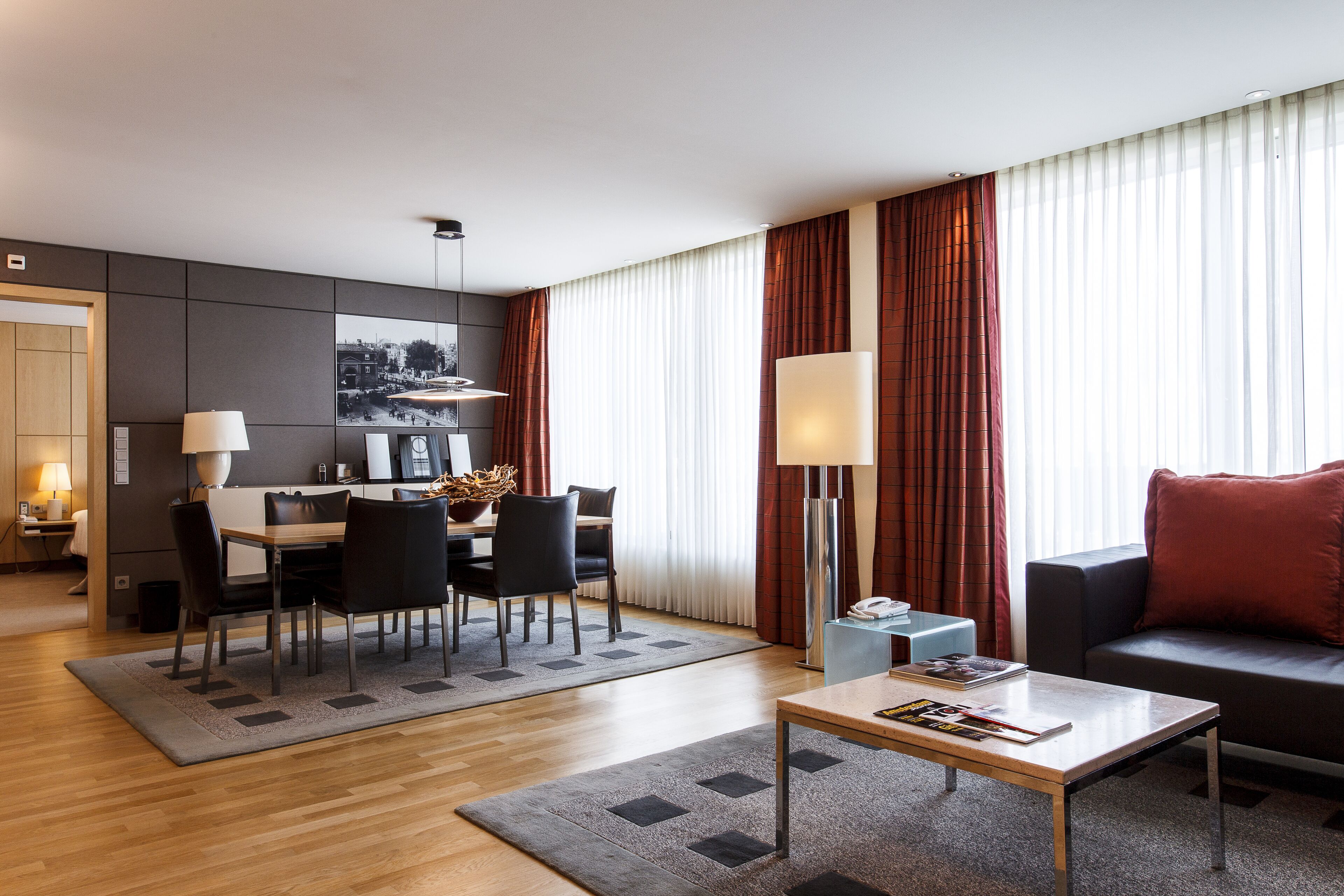 Quarto de hotel moderno com área de jantar, sofá, mesa de centro e uma grande janela com cortinas.