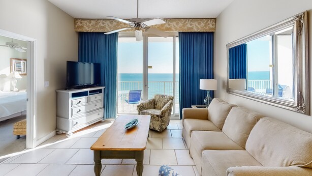 Quarto de hotel com varanda, televisão, sofá, mesa de centro e poltrona estampada.