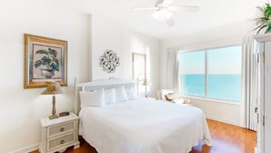 Quarto com uma cama grande, uma mesa de cabeceira com luminária, um objeto decorativo na parede e vista para o mar através de uma janela.