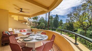 Uma sala de jantar com mesa redonda posta para quatro pessoas, rodeada por cadeiras de vime, com varanda e vista para um jardim e árvores.