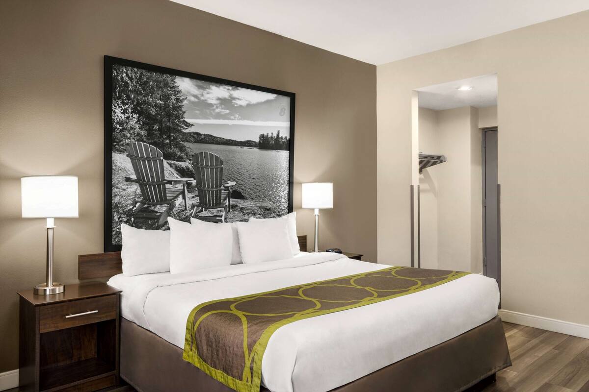 Quarto de hotel com uma cama grande, duas luminárias de cabeceira, um quadro com uma paisagem de lago e uma porta à direita.