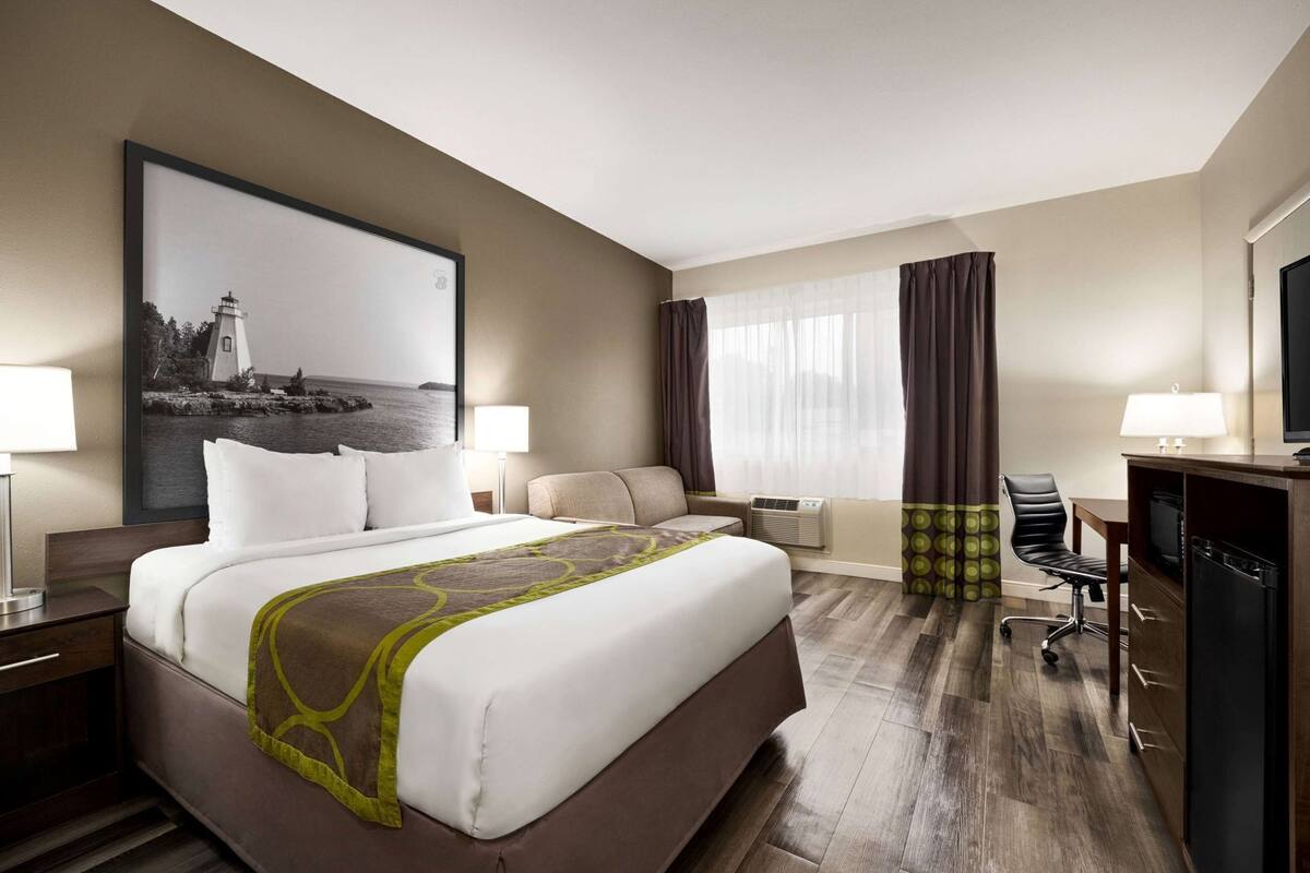Quarto de hotel com uma cama grande, uma escrivaninha com cadeira, uma televisão e uma janela com cortinas.