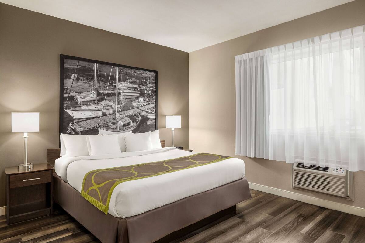 Quarto de hotel com uma cama grande, duas luminárias de cabeceira, uma janela com cortinas leves e um ar condicionado fixado na parede.