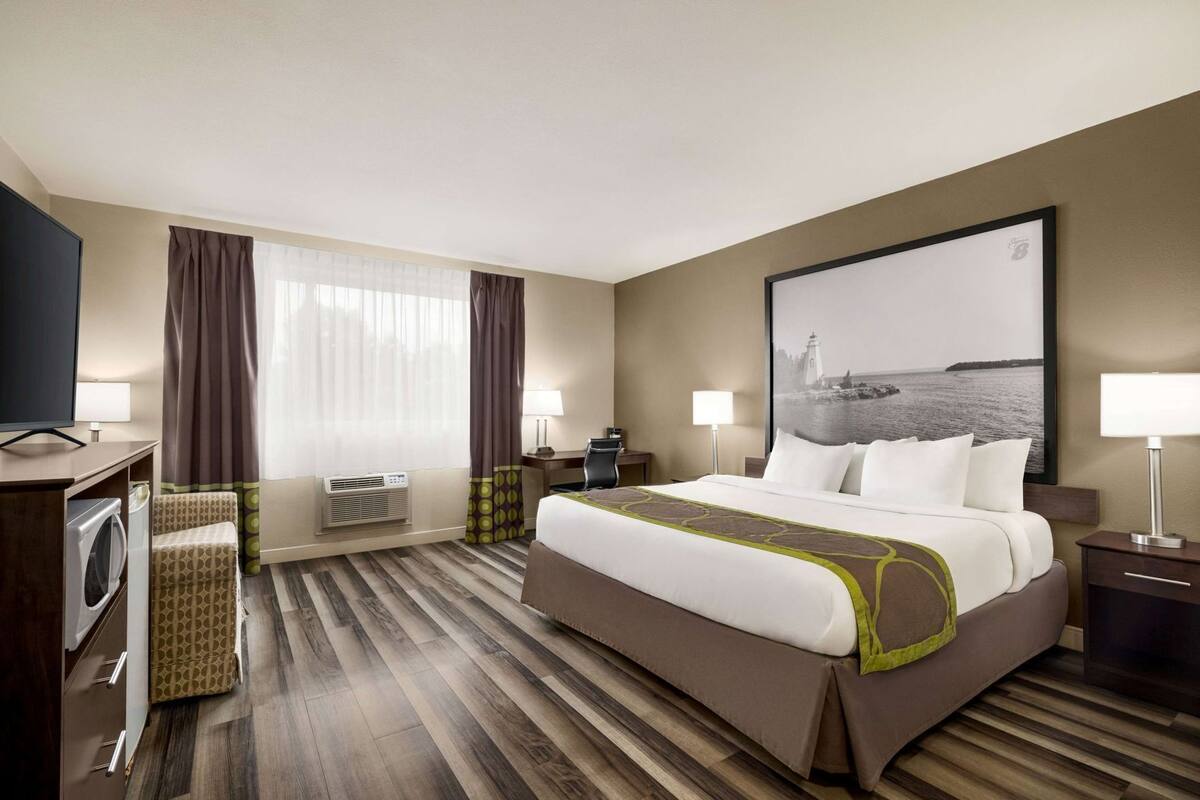 Quarto de hotel com cama, escrivaninha, cadeira, televisão, janela com cortinas e um quadro na parede.