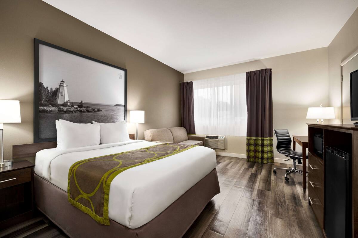 Quarto de hotel com uma cama grande, uma escrivaninha, uma cadeira, uma televisão e uma janela com cortinas.