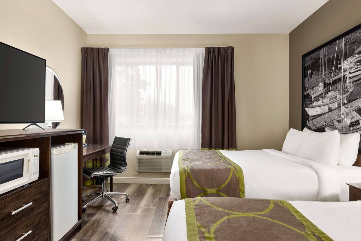 Quarto de hotel com cama, escrivaninha com micro-ondas, cadeira e uma janela grande com cortinas.