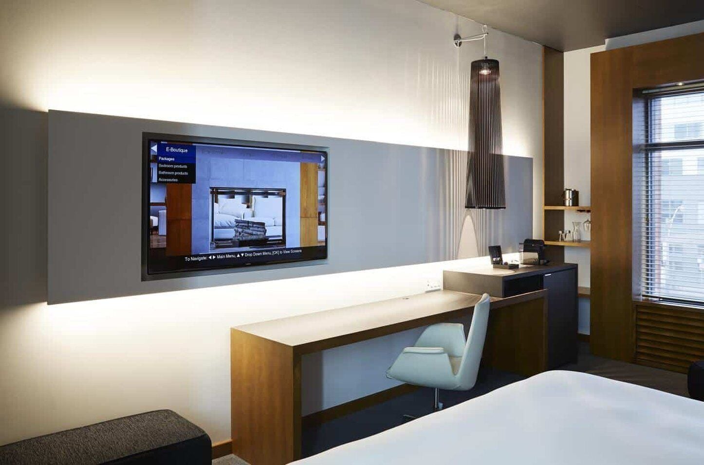 Quarto de hotel moderno com uma grande TV de tela plana na parede, uma escrivaninha de madeira, uma poltrona confortável, uma cama e uma janela com persianas.
