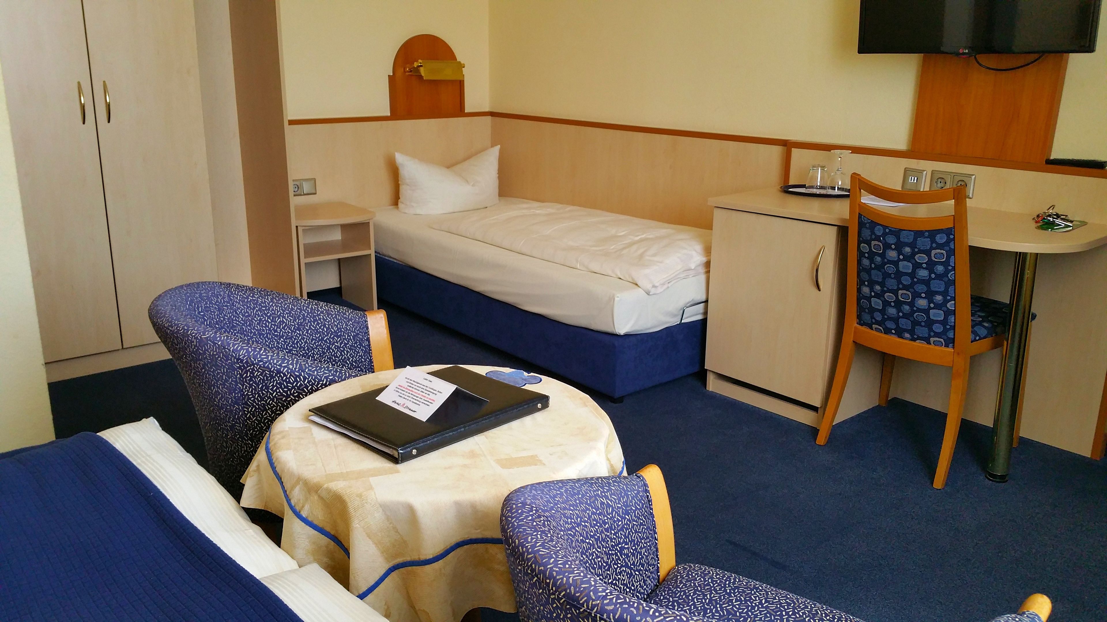 Quarto de hotel com uma cama de solteiro, uma pequena mesa com cadeira, um guarda-roupa e uma televisão.