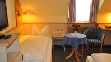 Quarto de hotel com uma cama, uma escrivaninha com um controle remoto, uma pequena mesa com uma placa, uma cadeira e uma janela com cortinas.