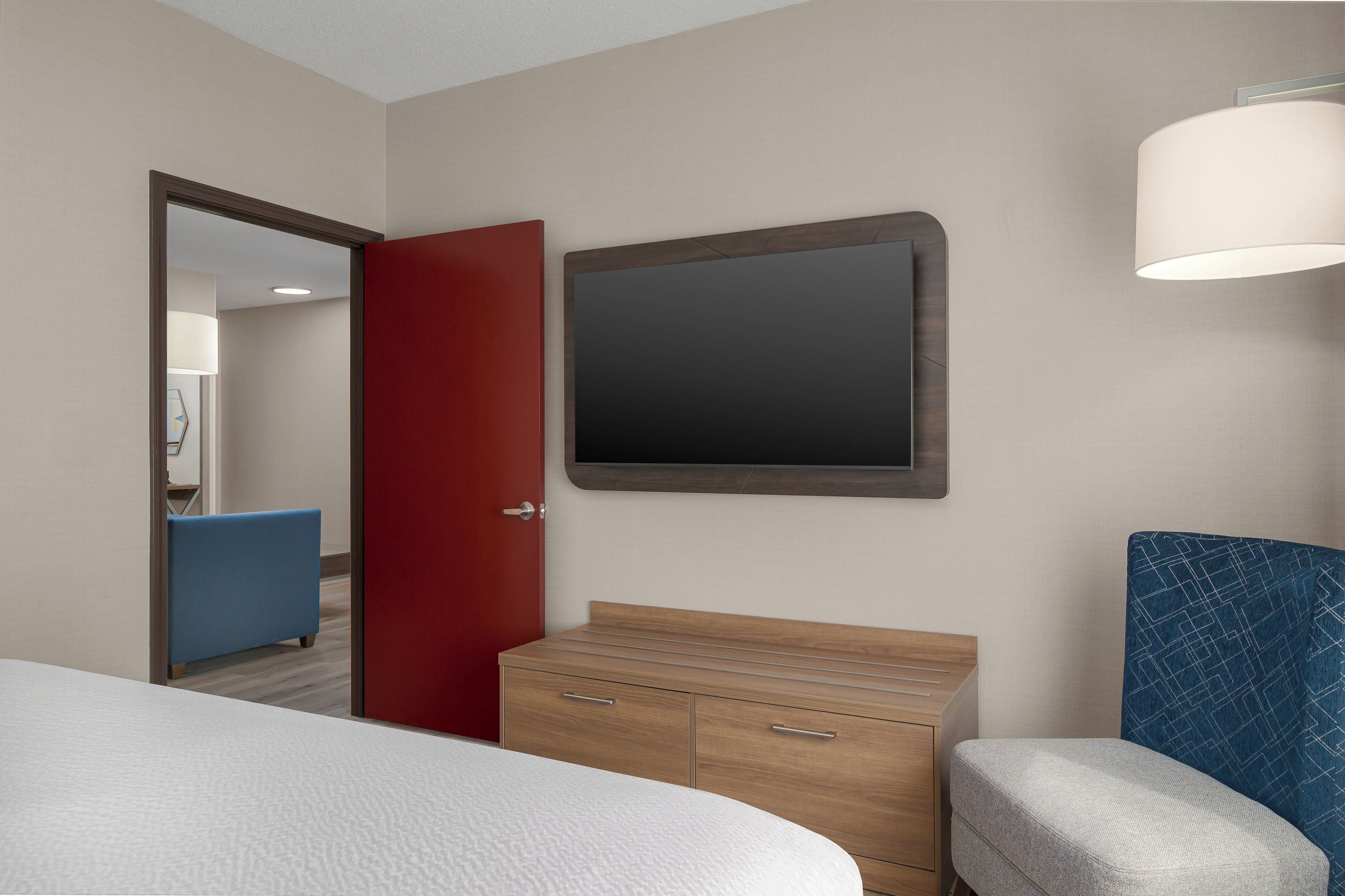 Quarto de hotel com cama, escrivaninha, cadeira, guarda-roupa, espelho e televisão.