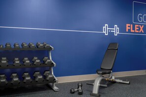 Academia com suporte de halteres fixado na parede, um banco e um tapete de ginástica.