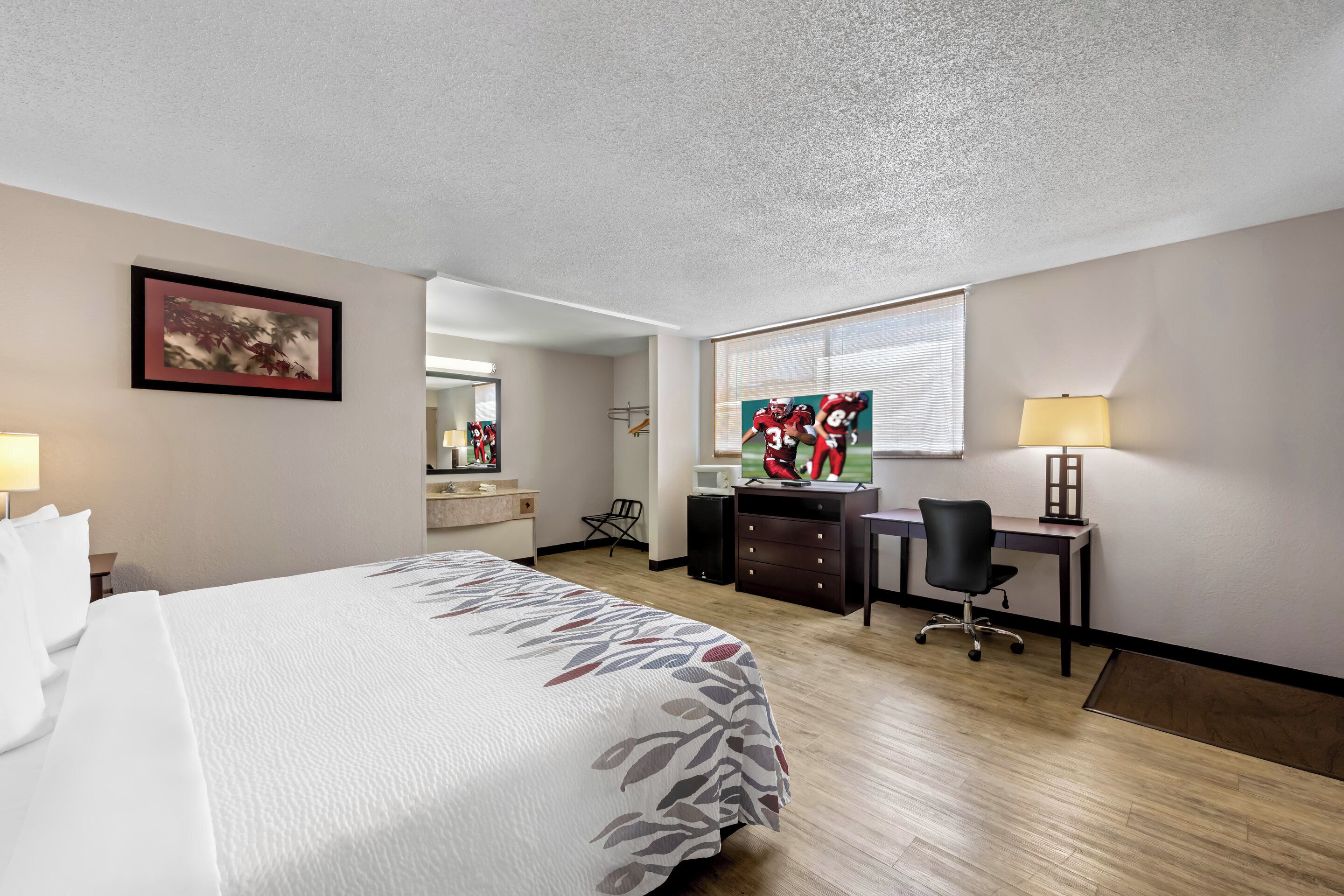 Quarto de hotel com uma cama grande, escrivaninha, cadeira, televisão e janela com persianas.