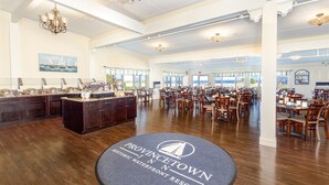 Uma sala de jantar ampla com várias mesas e cadeiras, um bar central e vista para o exterior.