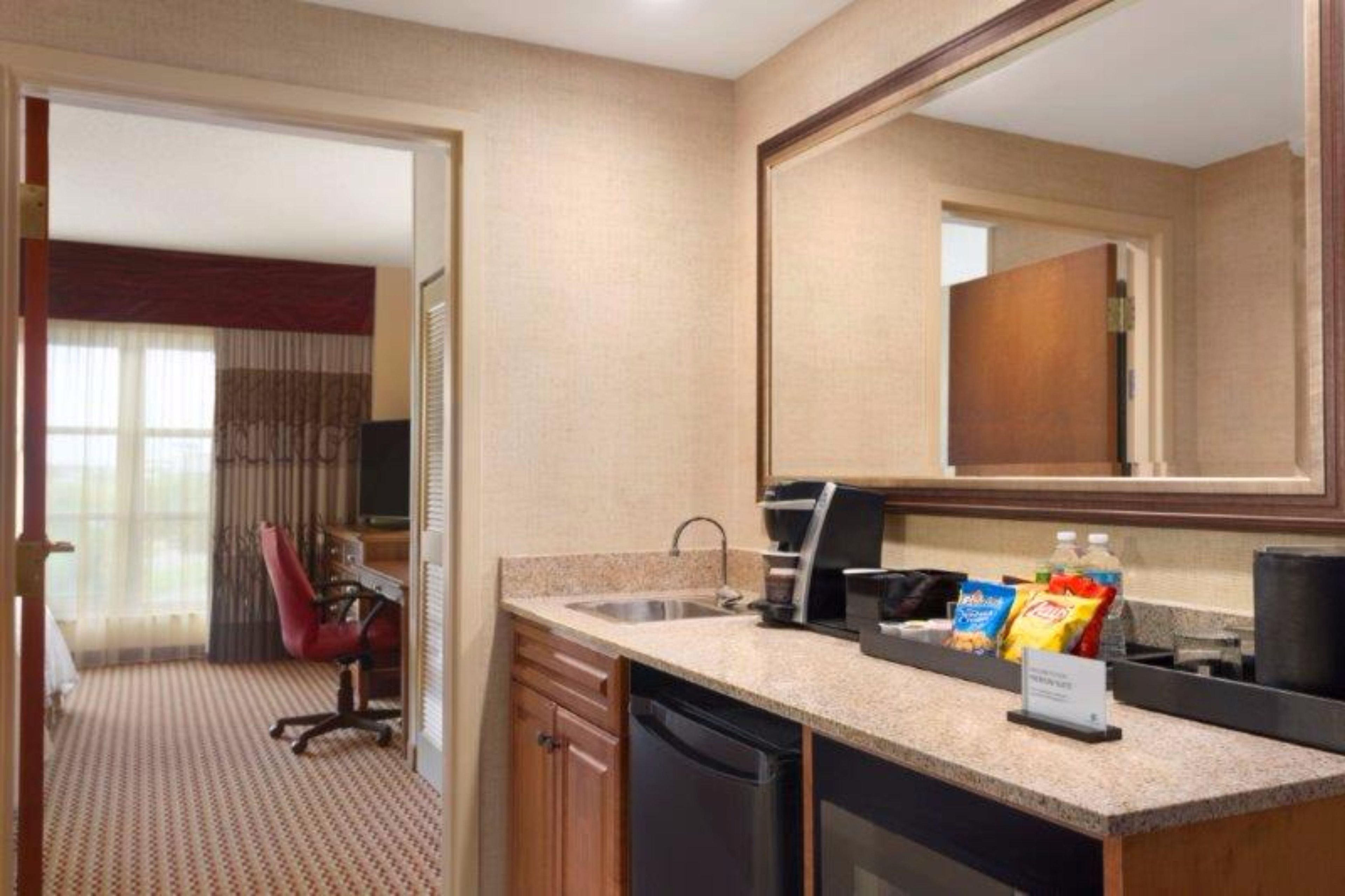 Quarto de hotel com cozinha compacta, área de refeições com uma cadeira vermelha, um espelho grande e vista para o corredor.