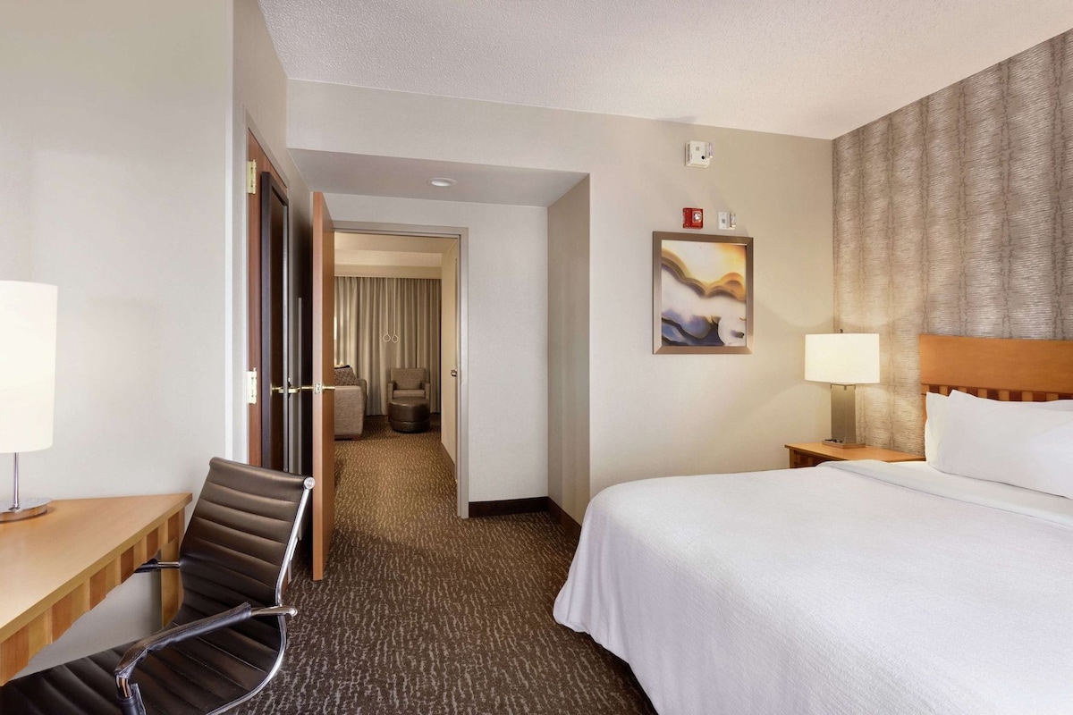 Quarto de hotel com cama, mesa com cadeira, banheiro com chuveiro e corredor que dá para outro quarto.