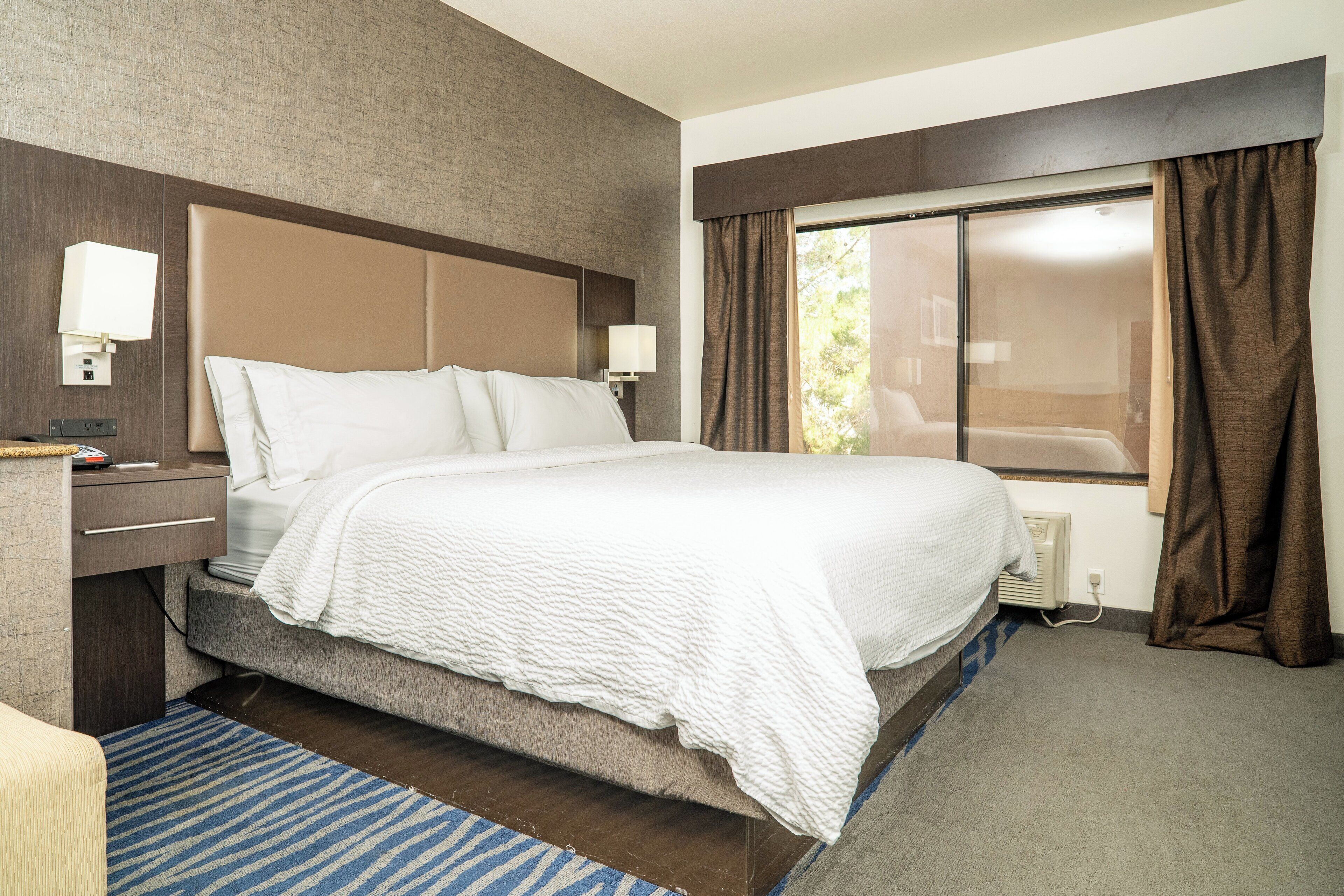 Quarto de hotel com uma cama grande, mesa de cabeceira, abajur e janela com cortinas.