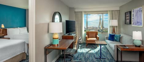 Quarto de hotel com cama, escrivaninha com luminária, cadeira, sofá, televisão e uma grande janela com vista para uma ponte.