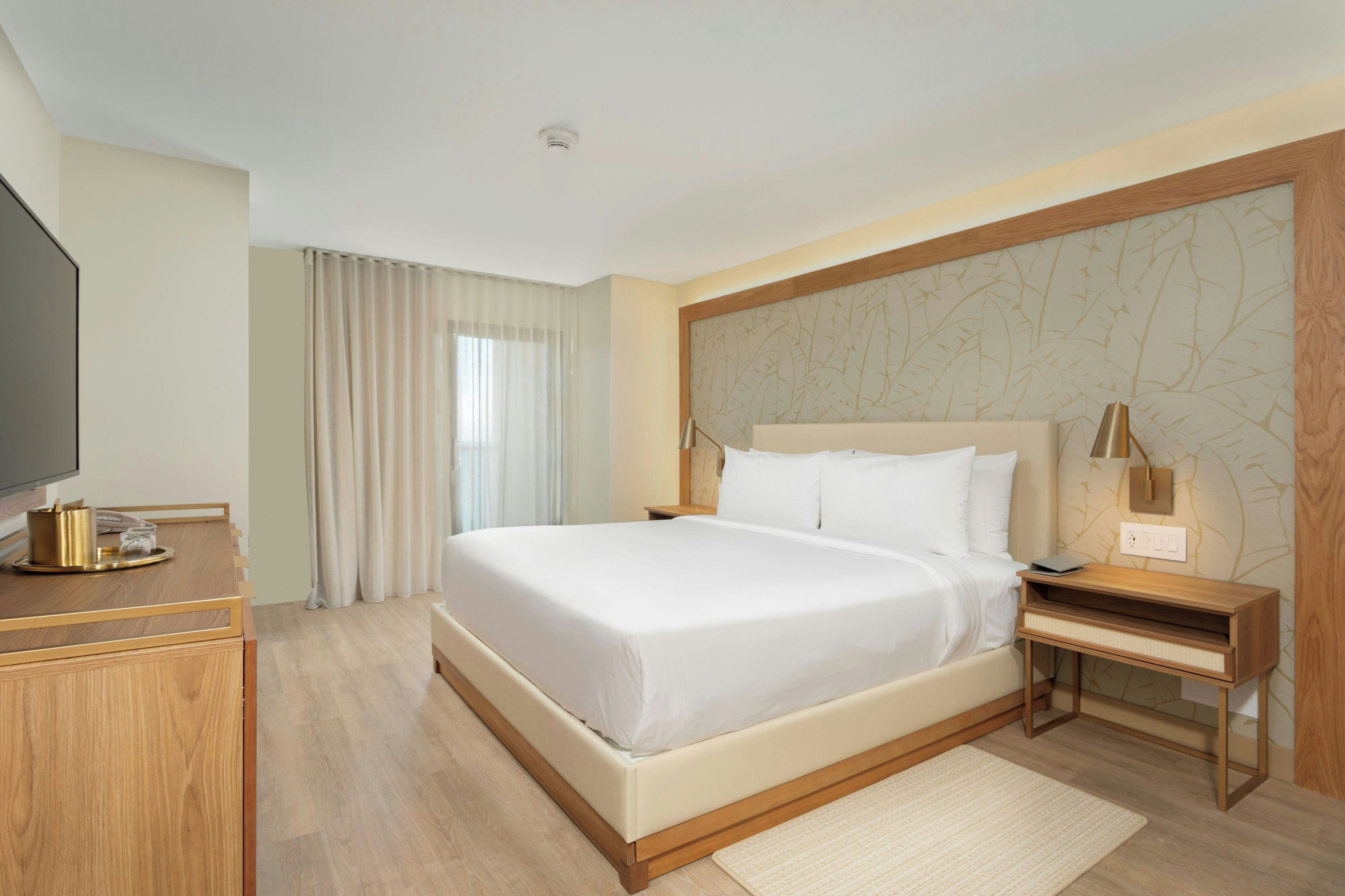 Quarto de hotel com uma cama grande, mesa de cabeceira, luminária e televisão sobre um suporte.