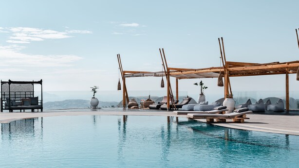 Uma área à beira da piscina com espreguiçadeiras, uma pérgola de madeira e um espaço de estar com vista para as montanhas.