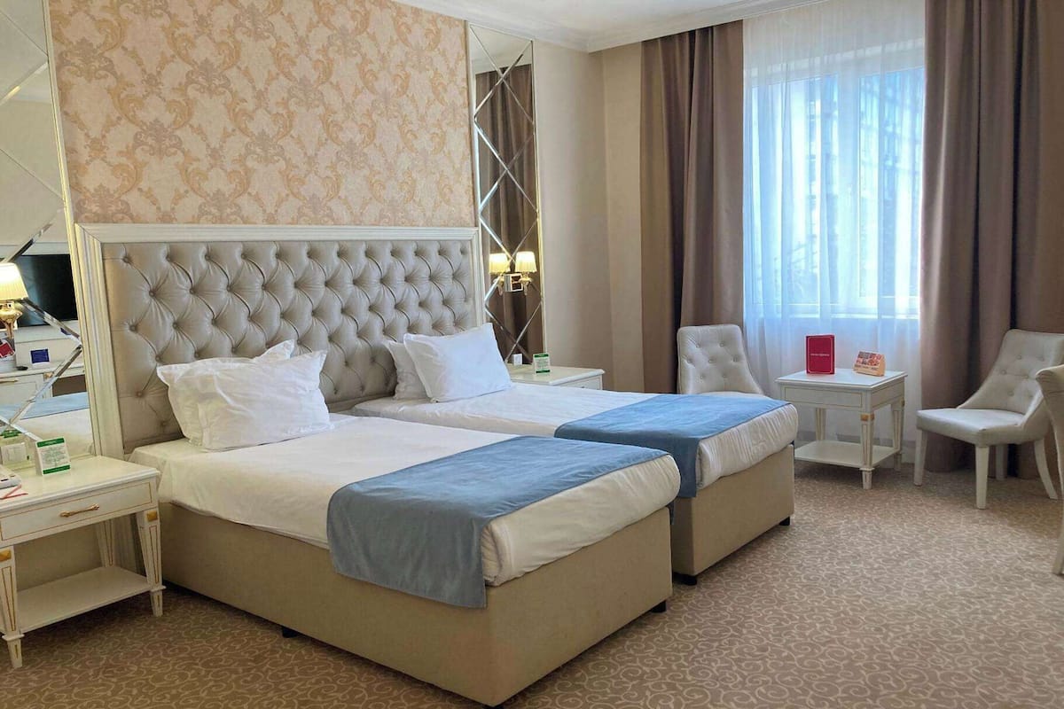 Quarto de hotel com duas camas, um espaço de estar com uma cadeira, uma mesinha e uma janela com cortinas.
